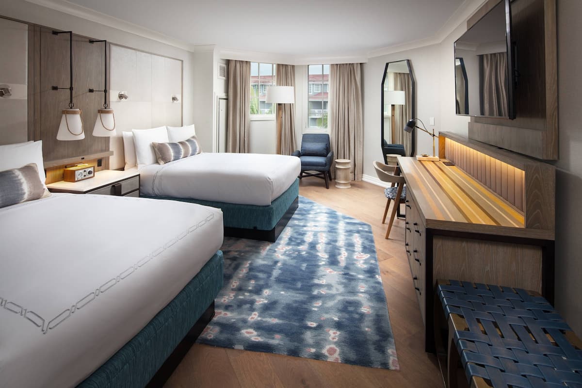 Quarto de hotel com duas camas, uma escrivaninha, uma cadeira, uma televisão e vista para edifícios.