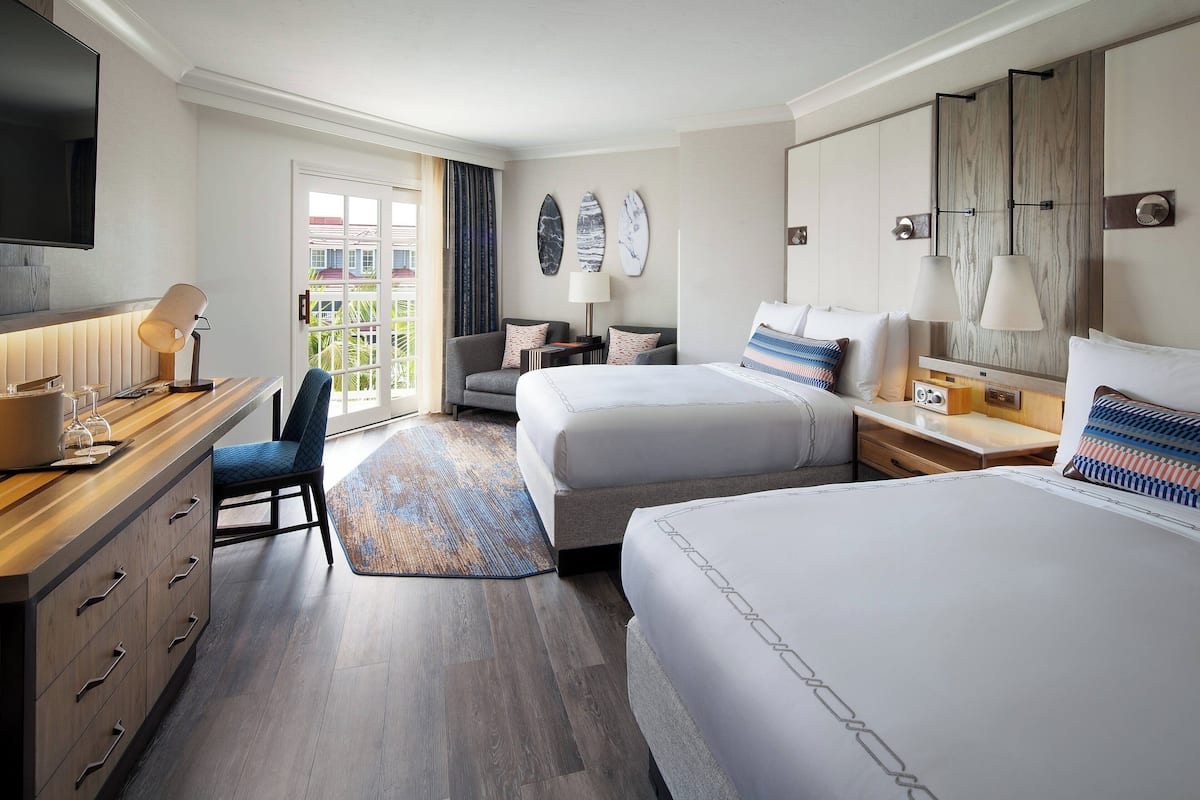 Quarto de hotel com uma cama grande, uma cama menor, uma mesa de trabalho com abajur, uma cadeira e vista para o exterior.