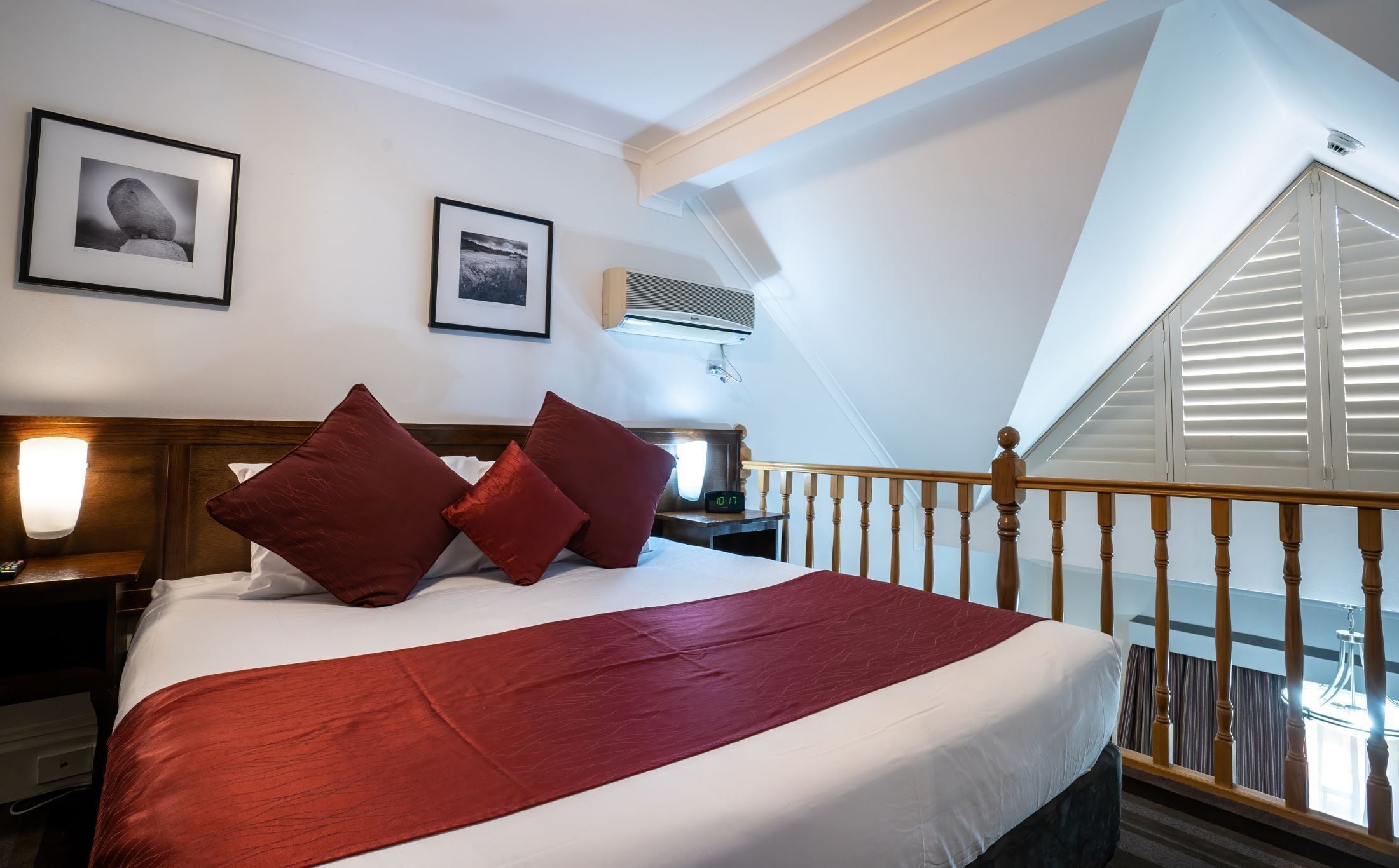 Quarto de hotel com uma cama grande, duas almofadas, uma mesa de cabeceira com abajur e duas imagens emolduradas na parede.