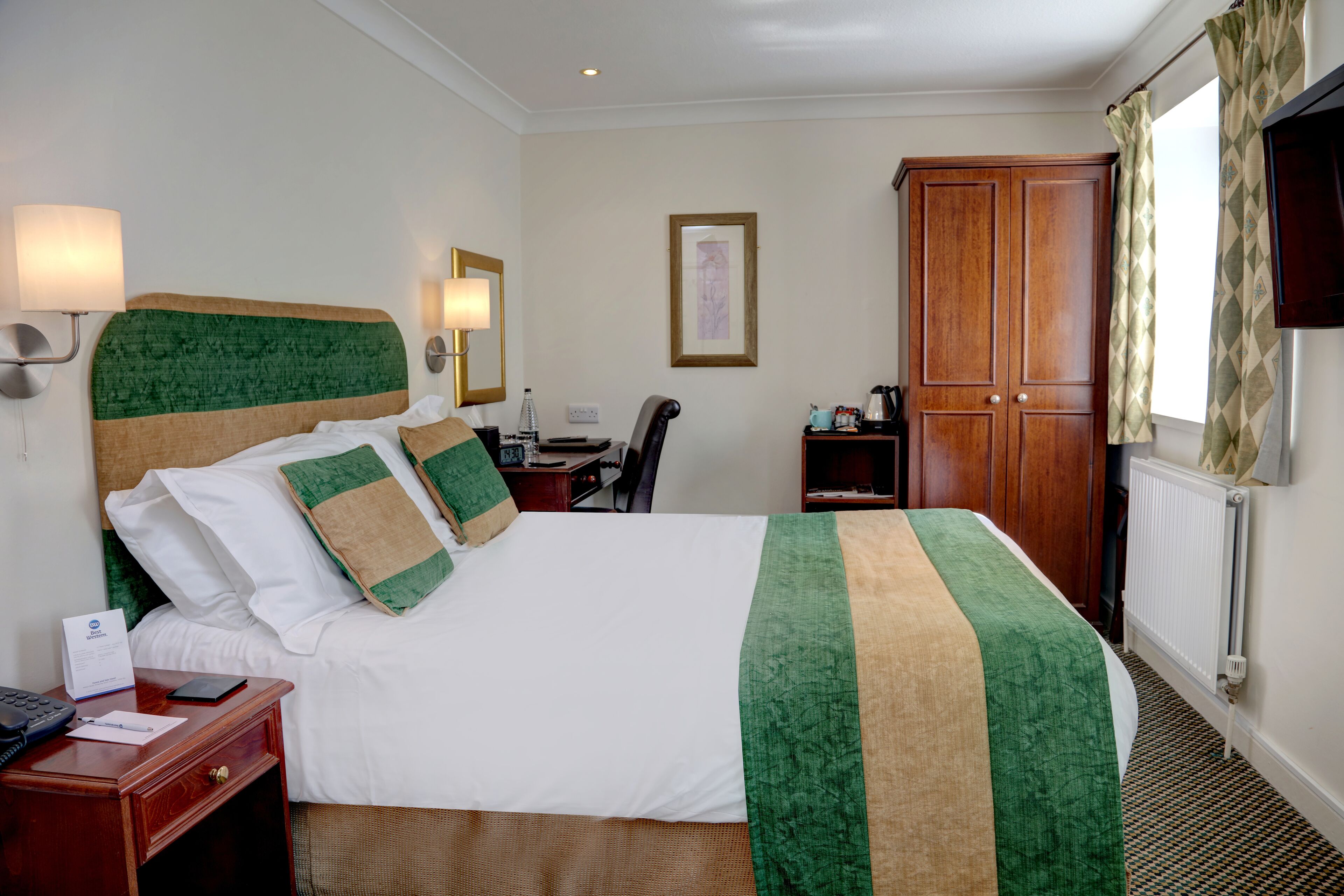 Quarto de hotel com uma cama grande, um guarda-roupa de madeira, uma escrivaninha com luminária e uma cadeira.