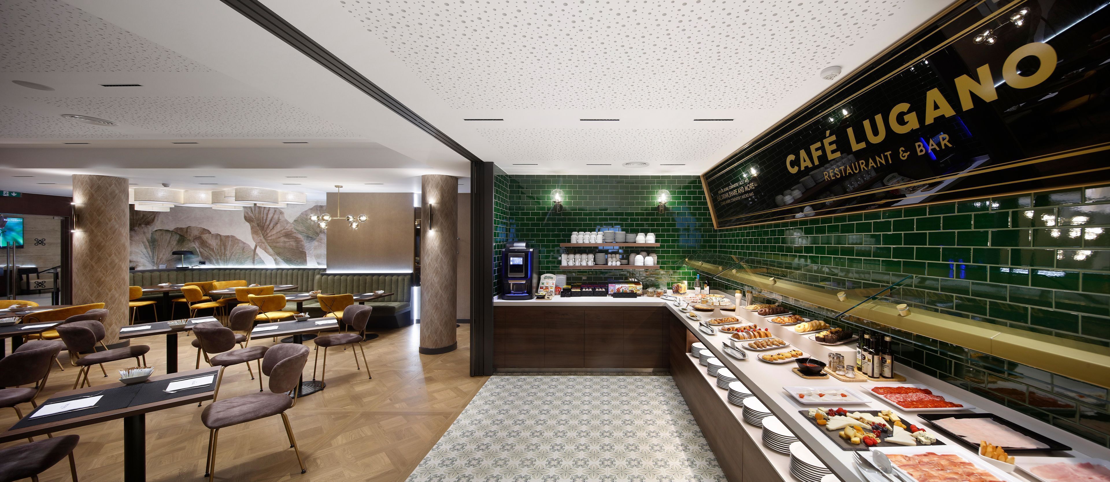Espaço café/lounge com assentos, balcão de buffet e parede revestida em azulejos verdes.