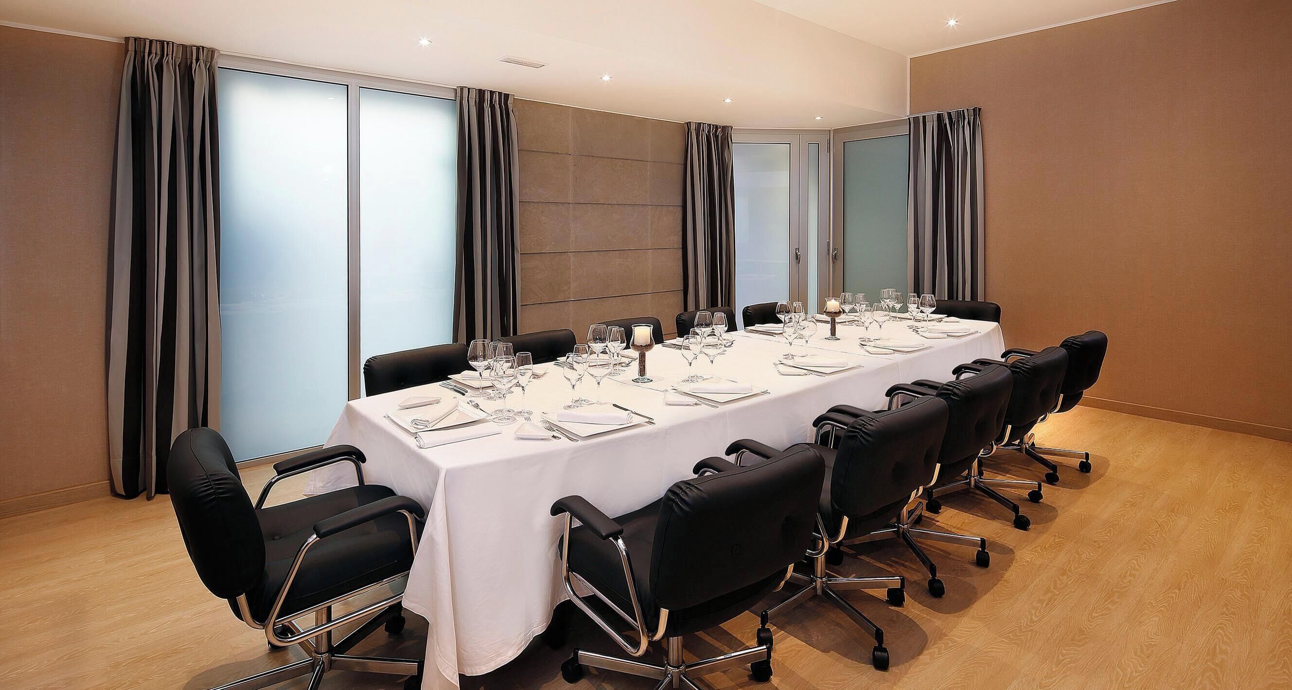 Sala de conferências com uma mesa comprida preparada para uma reunião, equipada com cadeiras e mesas, e grandes janelas com cortinas.