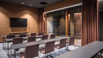 Uma sala de conferência com mesas longas e cadeiras, uma tela grande e um painel de vidro com cortinas.