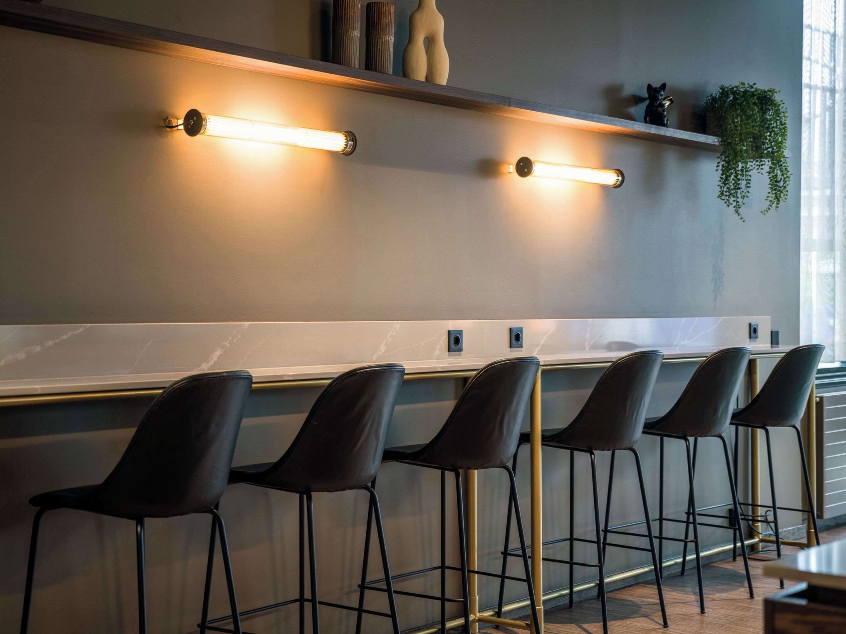 Uma área de bar com quatro banquetas, duas luzes fixadas na parede e uma prateleira com objetos decorativos.