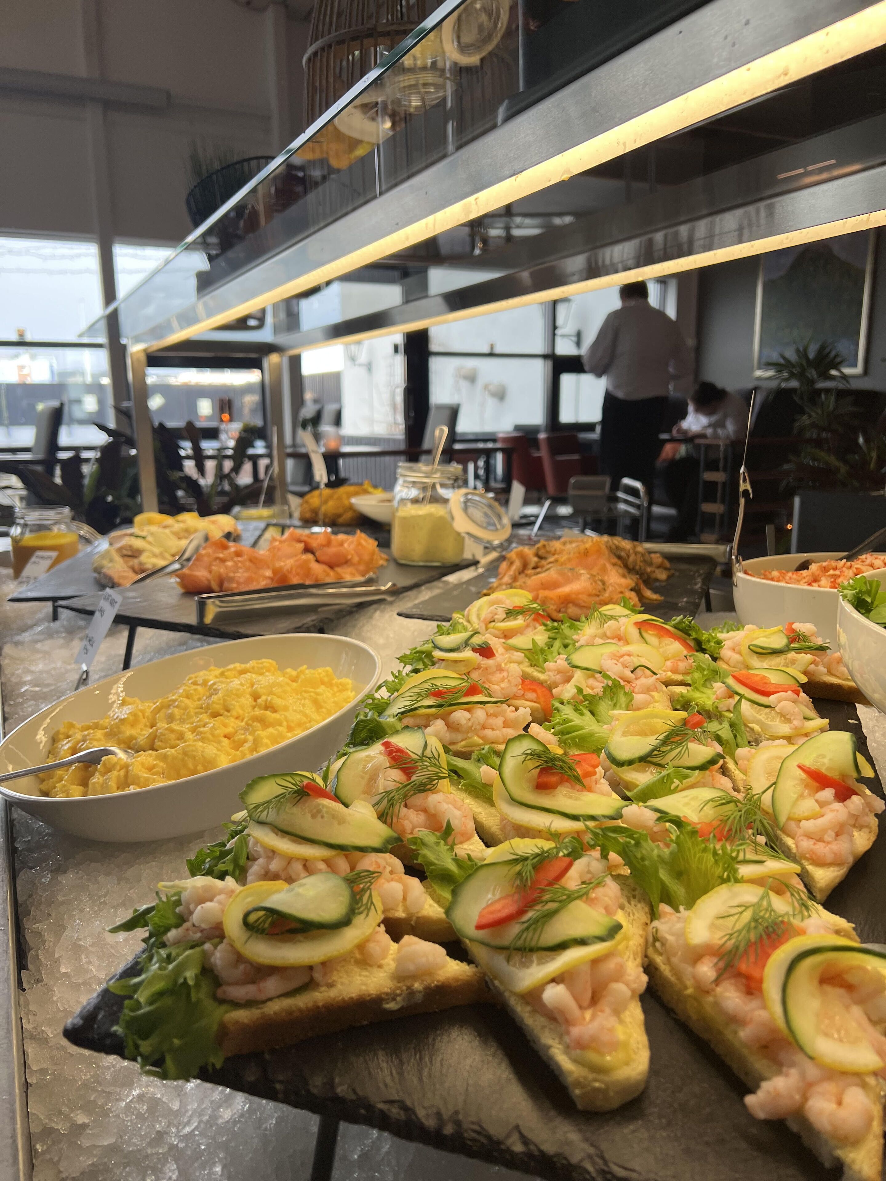 Buffet com diversos pratos, incluindo camarões no pão, ovos mexidos e saladas variadas.