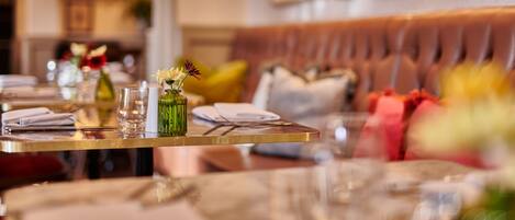 Uma sala de jantar com uma mesa posta para dois, um vaso verde com flores e quadros nas paredes.