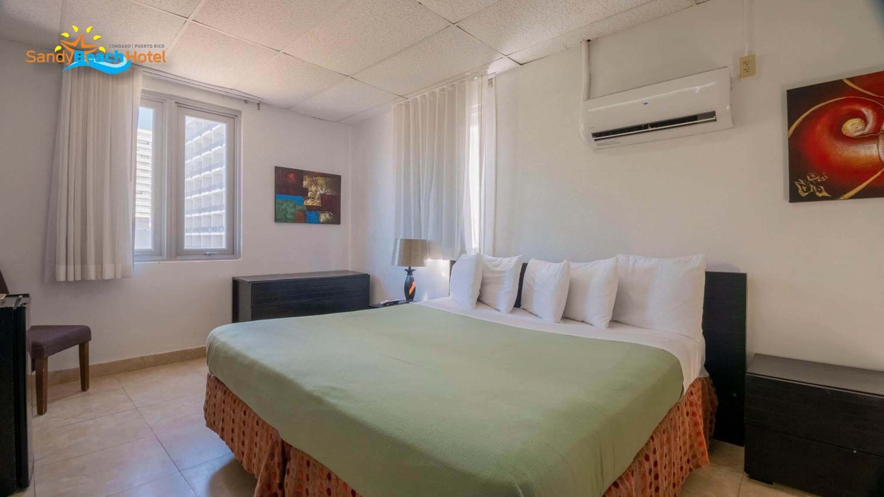 Quarto de hotel com cama, escrivaninha, cadeira, ar condicionado e um quadro na parede.