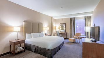 Quarto de hotel com uma cama grande, luminárias de cabeceira, escrivaninha com cadeira, televisão e janela com cortinas.