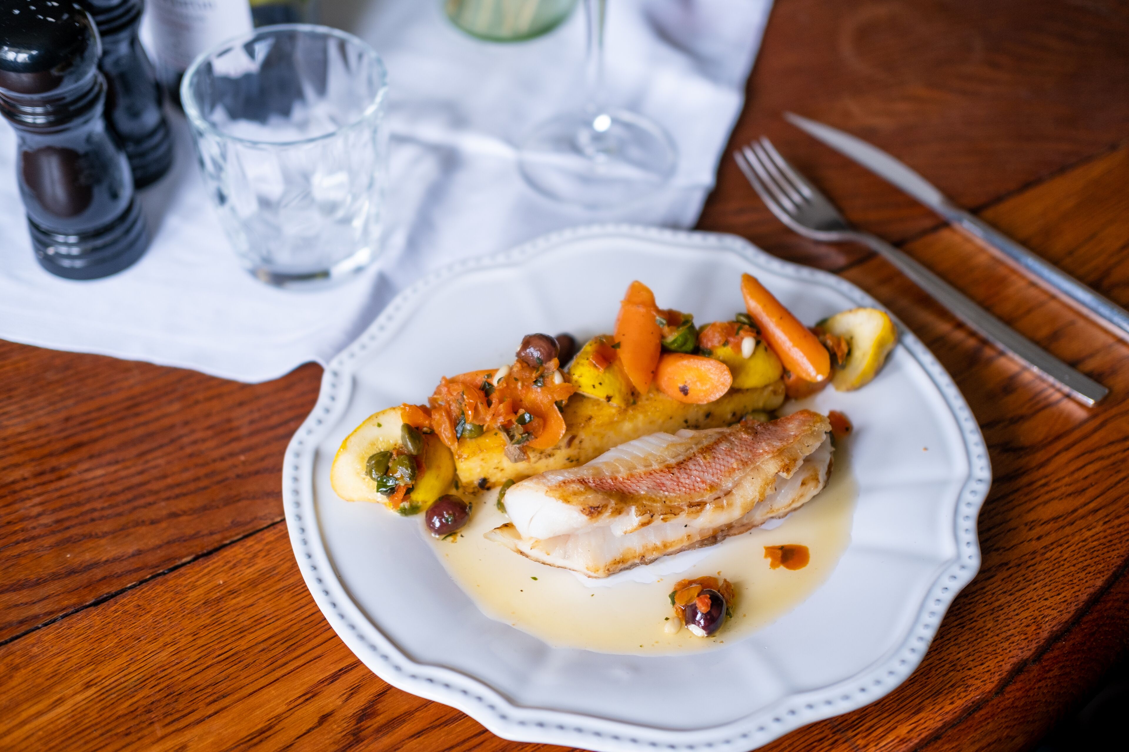 Um prato branco com um pedaço de peixe, legumes e molho sobre uma mesa de madeira.