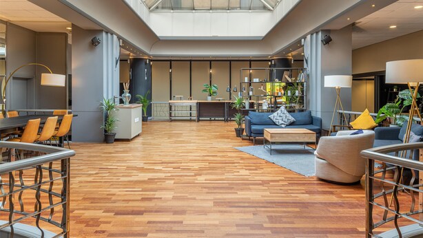 Um hall amplo com piso de madeira, áreas de estar, um balcão de recepção e uma cozinha ao fundo.