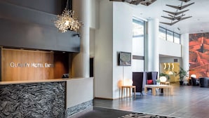 Uma área de recepção de hotel com design moderno, apresentando um balcão de atendimento, área de estar e um grande mural abstrato na parede.