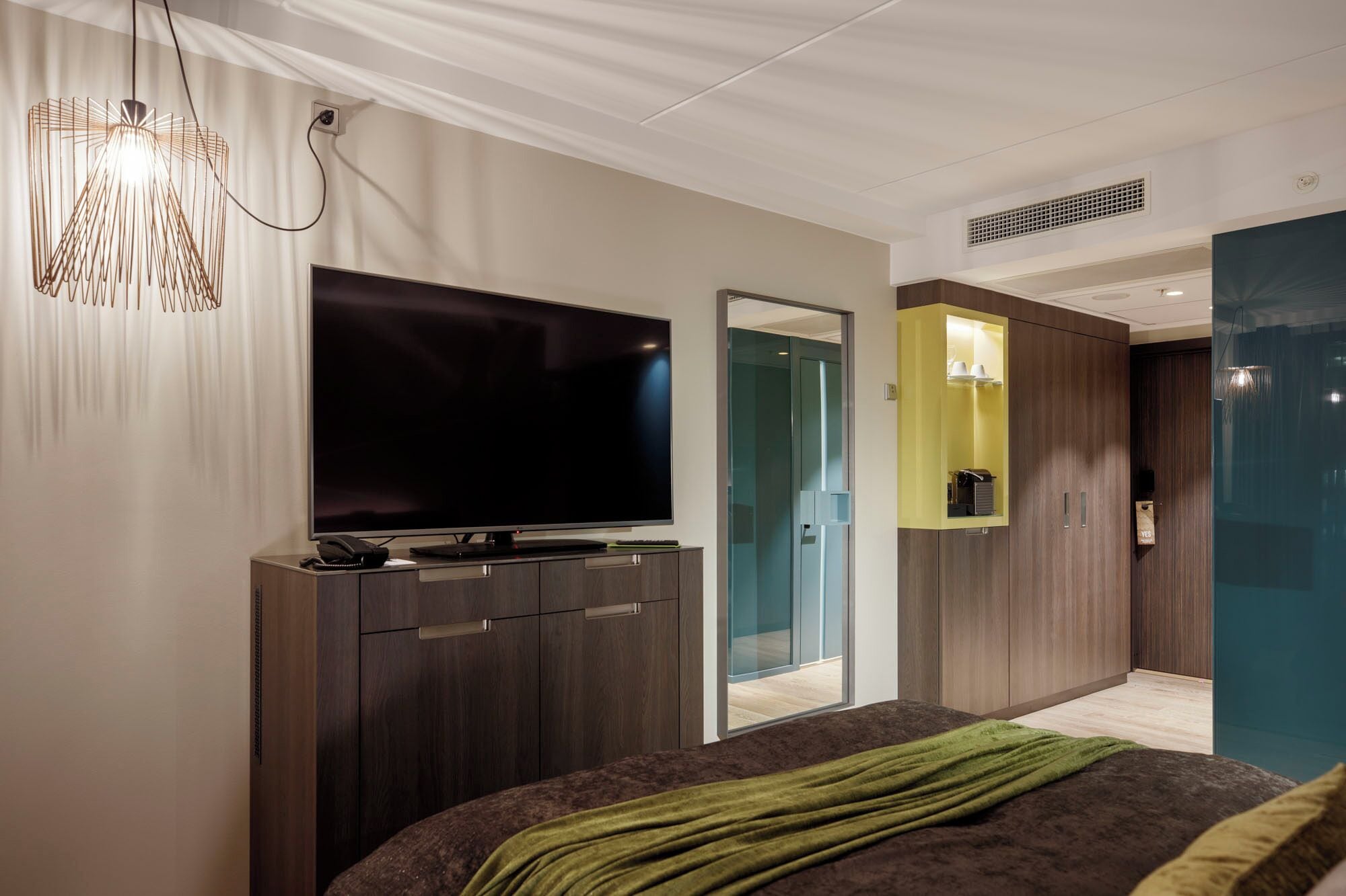 Quarto de hotel moderno com uma televisão de tela plana grande, um armário de madeira, um cobertor verde e um espelho com porta de vidro.