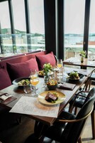 Restaurante com mesa de madeira posta para refeição, menu e vista da cidade através de grandes janelas.