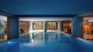 Piscina interior com água azul cristalina, rodeada por espreguiçadeiras e uma parede de pedra.