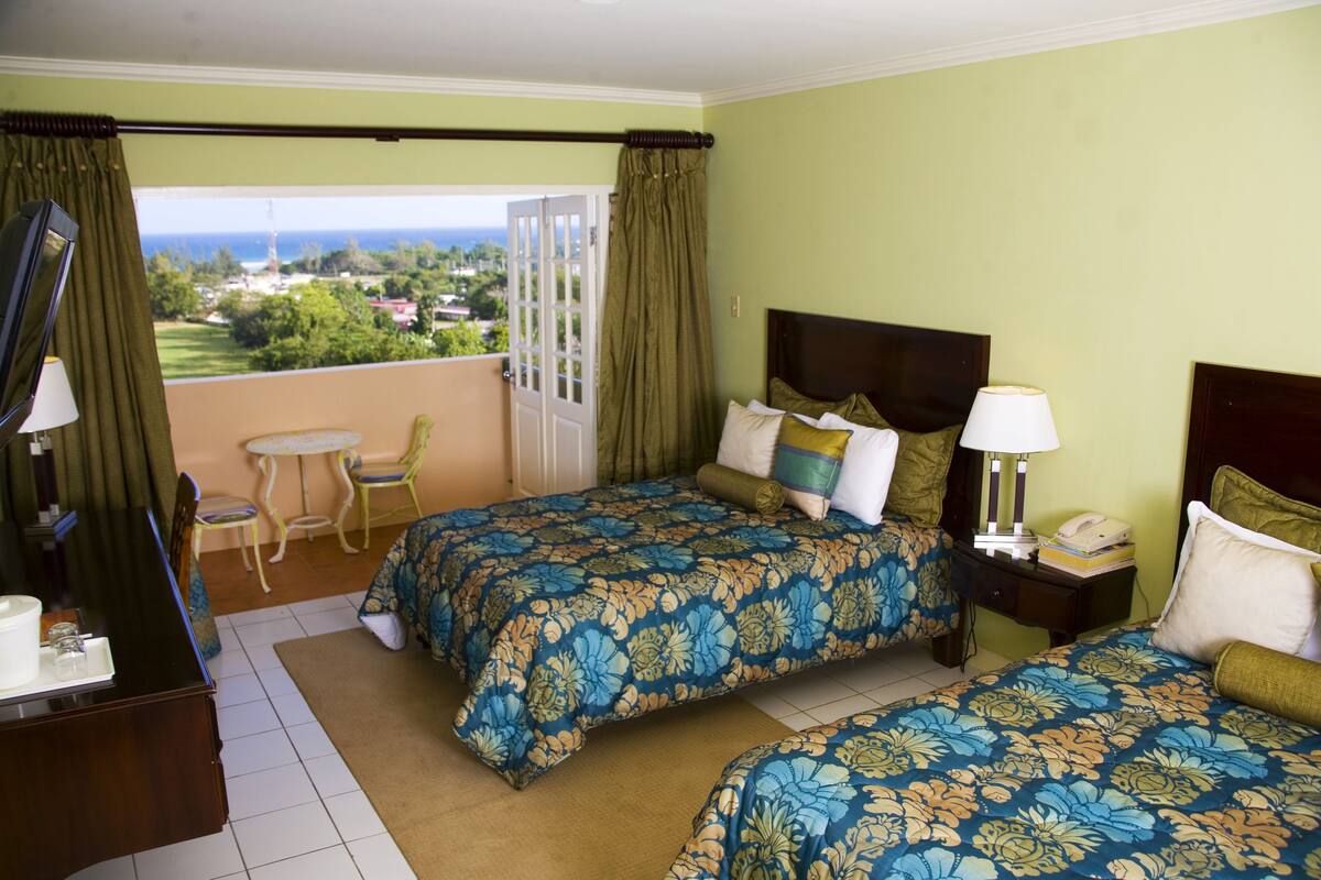Quarto de hotel com duas camas, uma varanda pequena com mesa e cadeiras, e vista para a paisagem.
