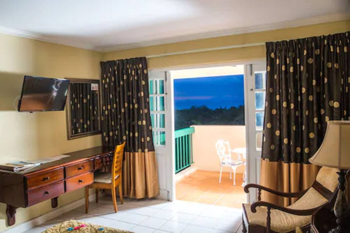 Quarto com escrivaninha, cadeira, televisão e varanda com cortinas.