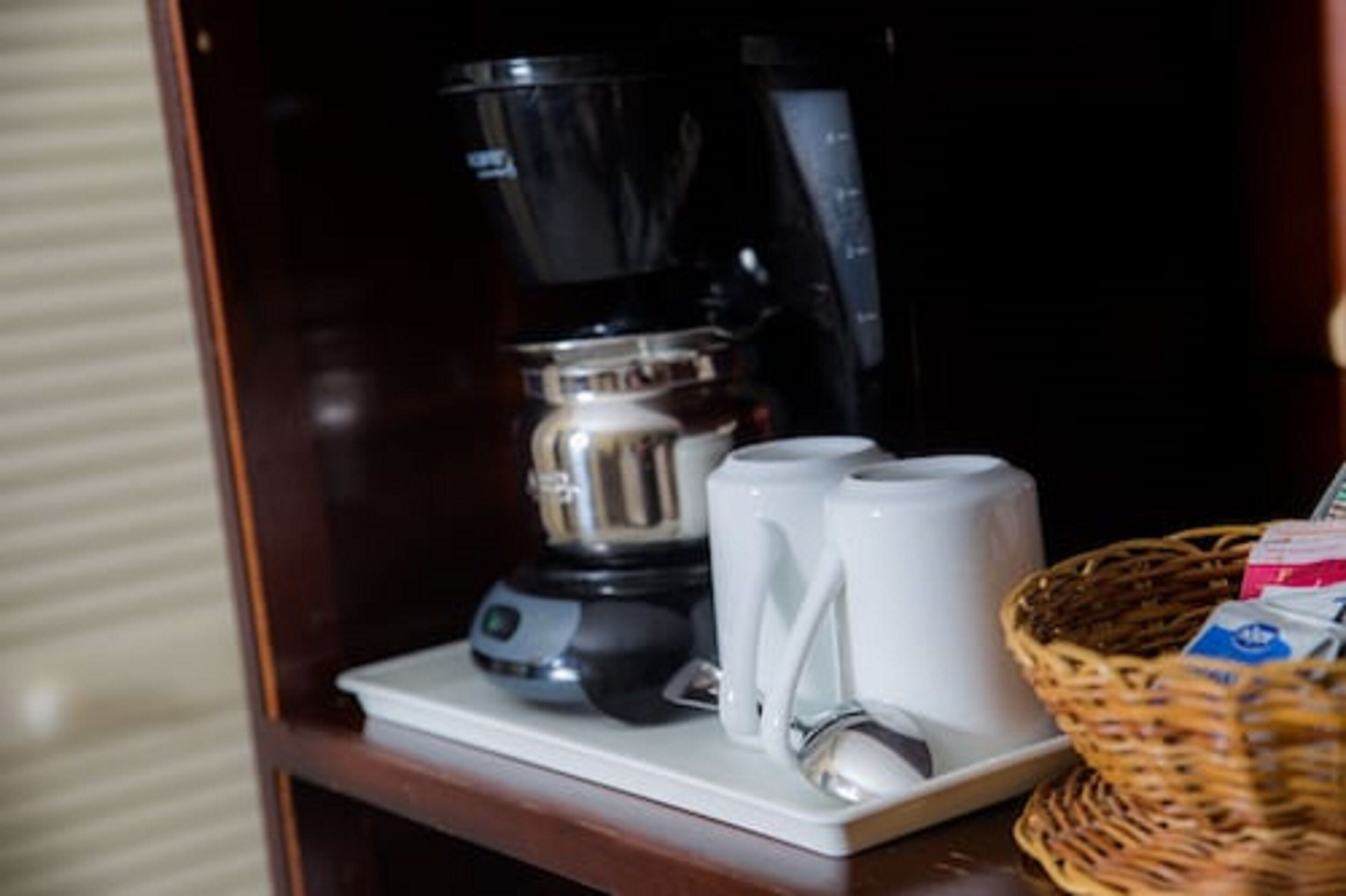 Máquina de café sobre um tabuleiro branco, com duas chávenas brancas e uma colher, uma cesta com objetos, e um aparelho de cor escura.