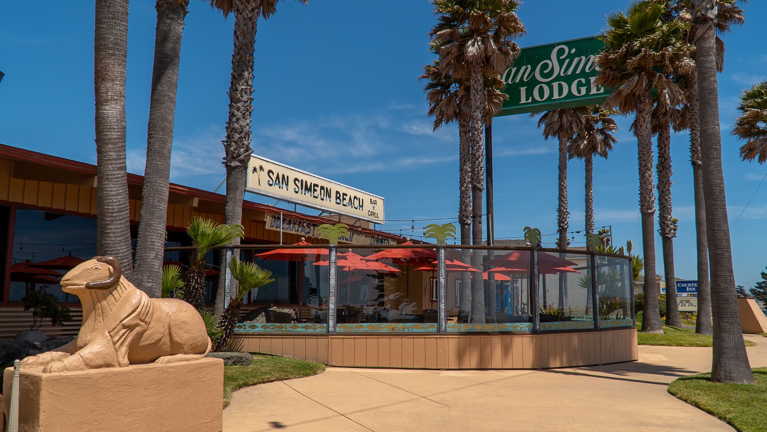 Um restaurante chamado 'San Simeon Beach Bar & Grill', com uma placa, palmeiras e uma estátua de um carneiro.