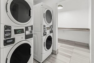 Lavanderia industrial com várias máquinas de lavar, painéis de controle e piso ladrilhado.