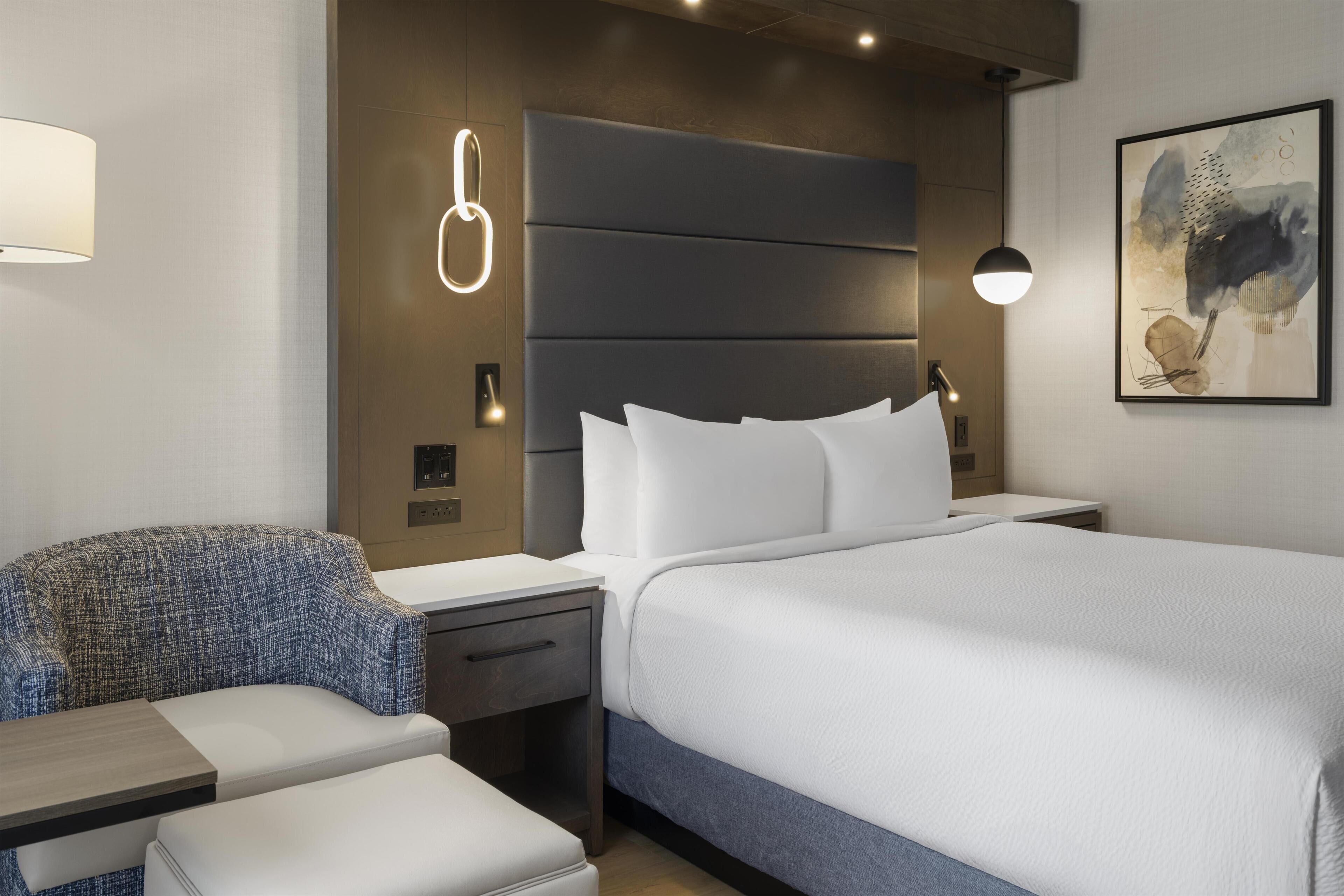 Quarto de hotel moderno com uma cama grande, uma escrivaninha, uma cadeira e uma mesinha de cabeceira.