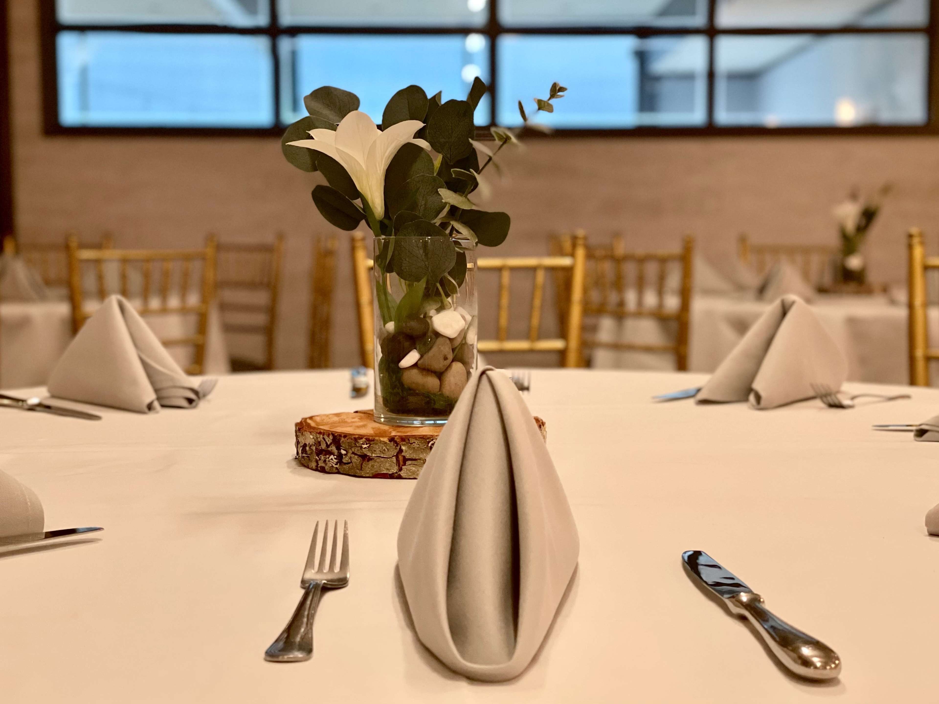 Uma mesa de jantar posta, com um vaso de flores brancas, uma toalha dobrada e talheres.