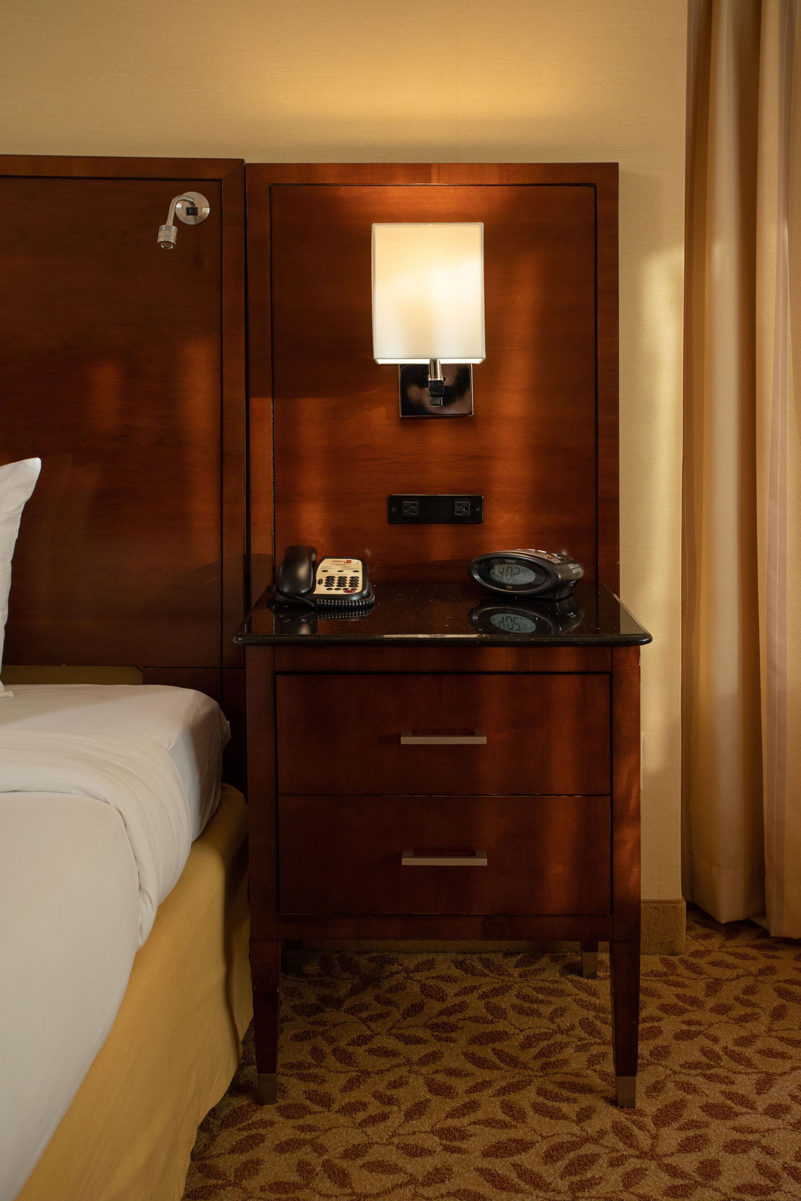 Quarto de hotel com uma cama, uma mesa de cabeceira de madeira com abajur, um telefone e um relógio.