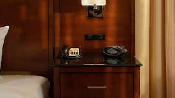 Quarto de hotel com uma cama, uma mesa de cabeceira de madeira com abajur, um telefone e um relógio.