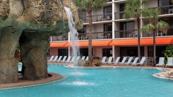 Piscina de hotel com cachoeira, cadeiras de descanso e um edifício com varandas.