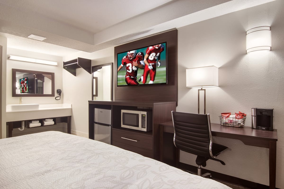 Quarto de hotel com cama, secretária com computador, televisão, micro-ondas, cadeira e luminária.