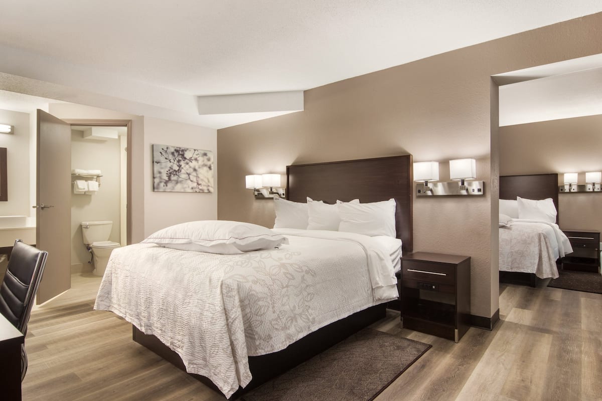 Quarto de hotel moderno com uma cama grande, mesas de cabeceira, uma escrivaninha e um banheiro visível por uma porta aberta.