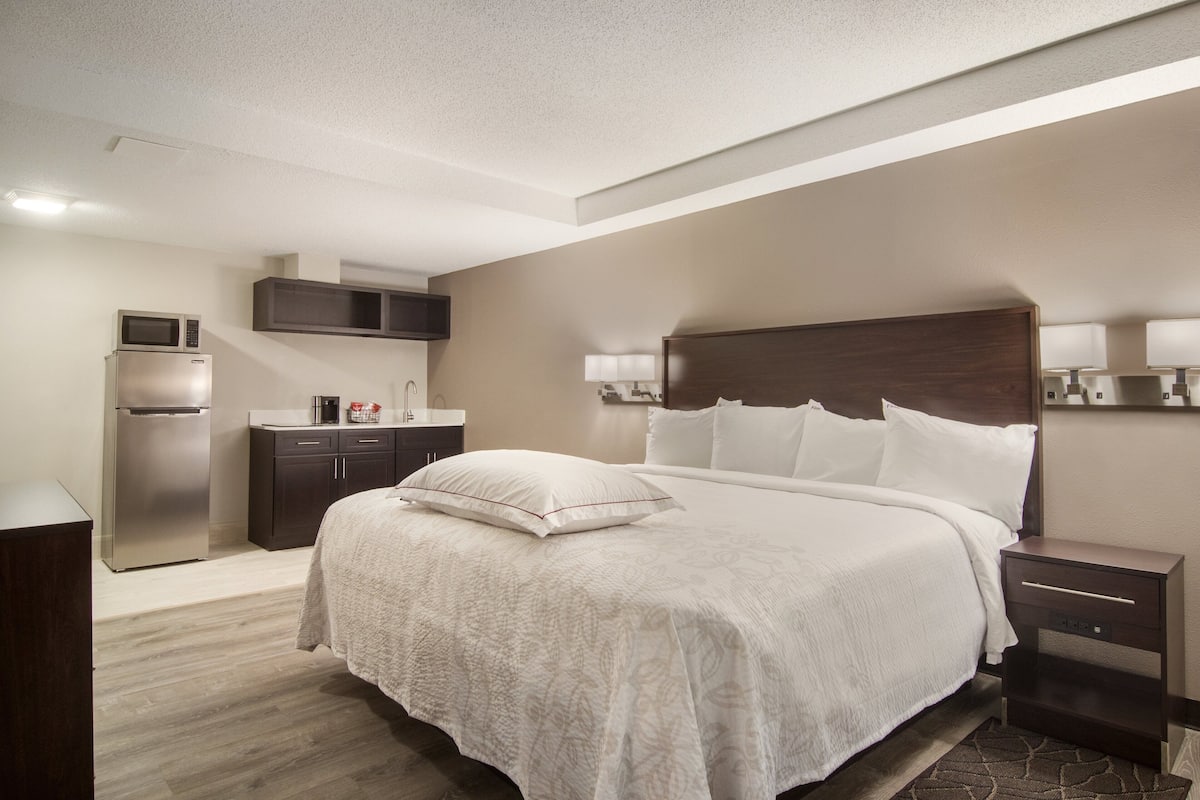 Quarto de hotel moderno com uma cama grande, micro-ondas embutido, frigorífico, uma pequena copa e uma mesa de cabeceira com uma luminária.