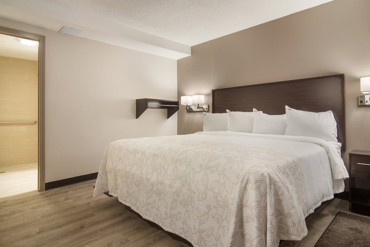 Quarto de hotel com uma cama grande, mesa de cabeceira, luminária na parede e piso de madeira.
