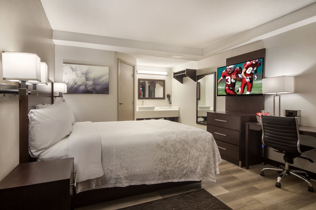 Quarto de hotel com cama, escrivaninha, cadeira, televisão e banheiro visível por uma porta aberta.