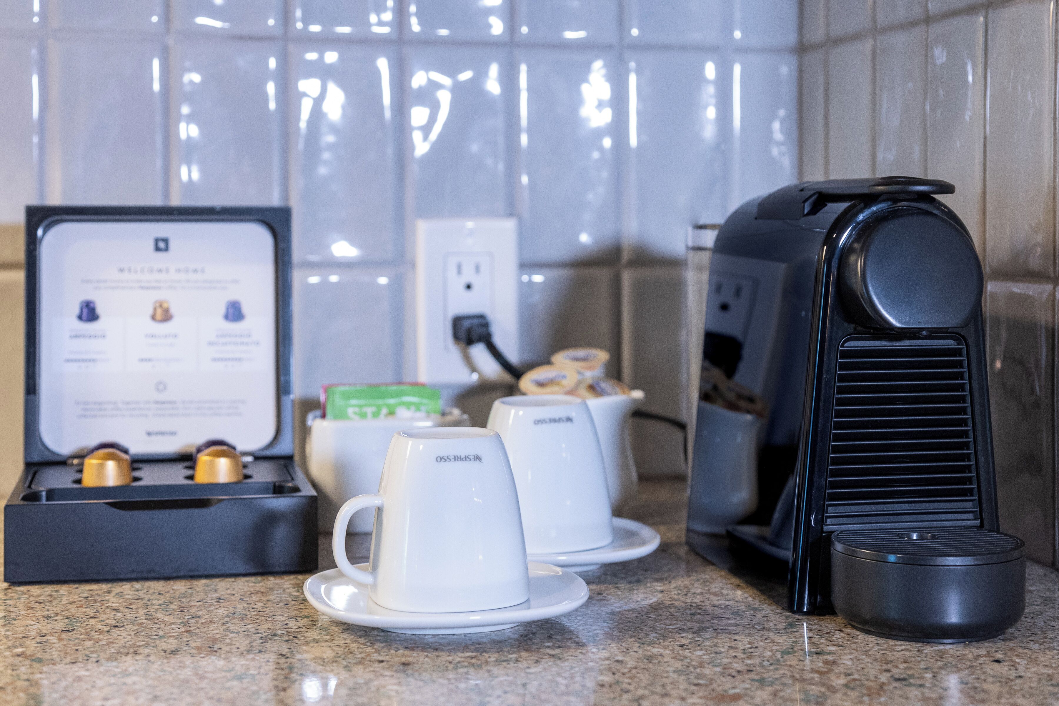 Uma estação de café com uma cafeteira, duas xícaras brancas e uma caixa de cápsulas de café.