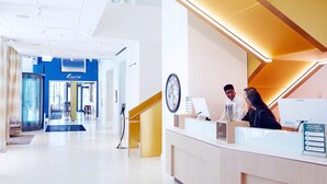 Uma área de recepção de hotel com design moderno, incluindo um balcão de atendimento, um relógio e uma placa de aviso com eventos futuros.