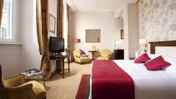 Quarto de hotel com uma cama grande, televisão, duas poltronas, uma mesa pequena e janela com cortinas.