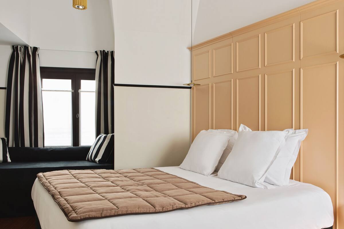 Uma cama bem arrumada com roupa de cama branca e um manta acolchoada marrom, um sofá escuro com almofadas listradas e uma janela com cortinas.