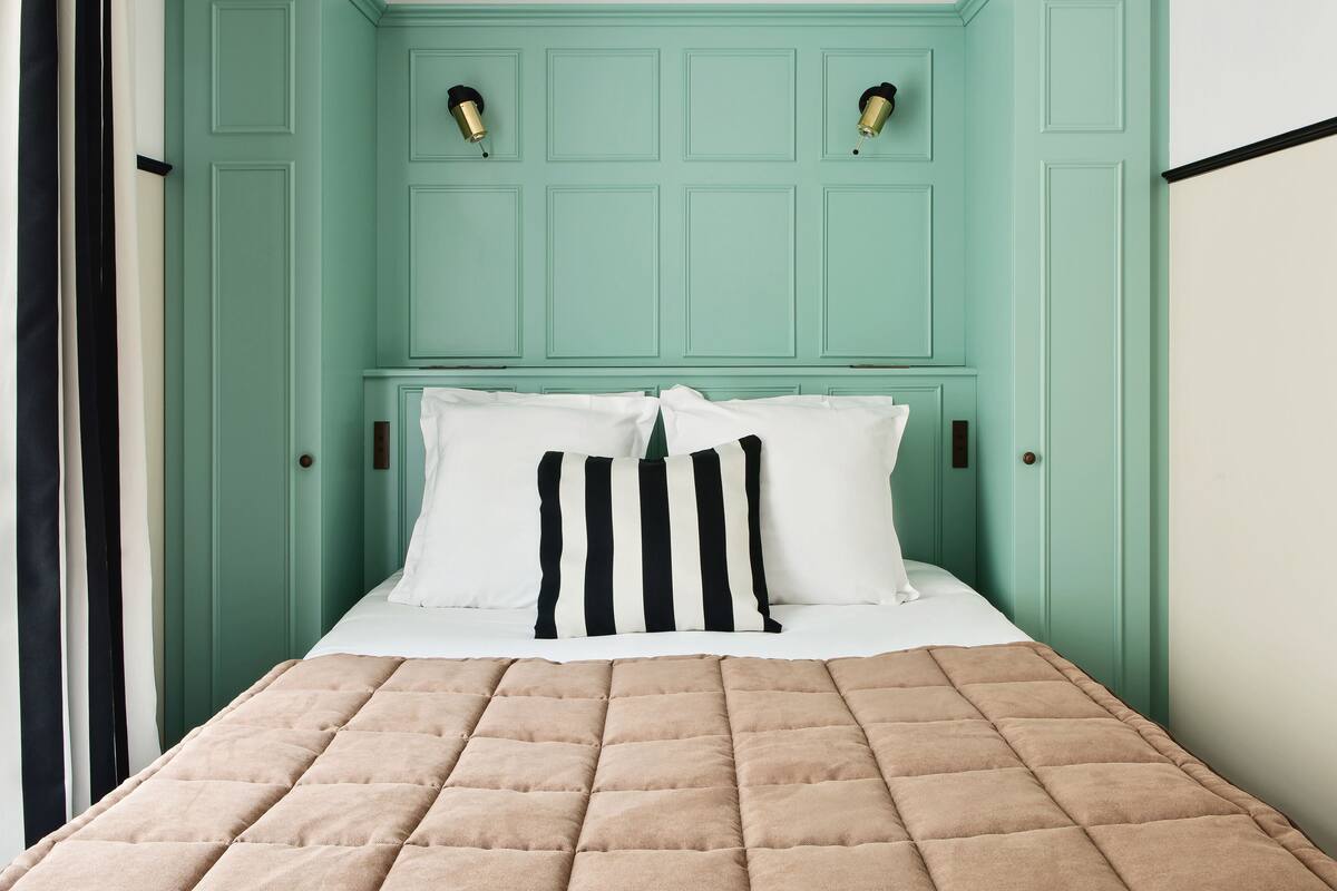 Cama com colcha bege acolchoada, almofadas brancas e uma almofada listrada em preto e branco. O quarto tem paredes verde-água e um guarda-roupa verde-água combinando, com duas luminárias.
