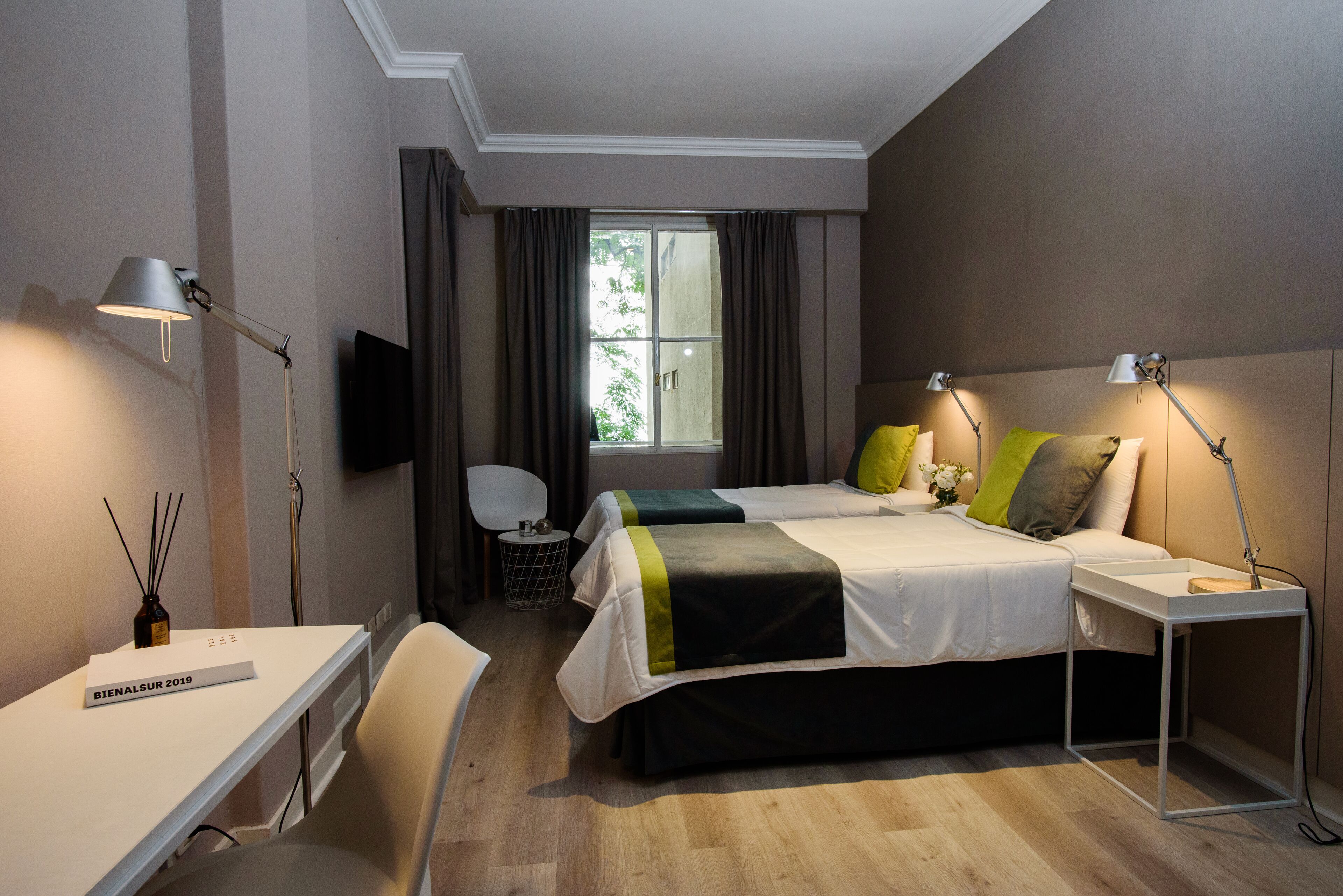 Quarto de hotel com duas camas, uma escrivaninha, uma cadeira, uma televisão e uma janela com cortinas.