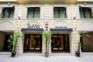 Entrada de hotel com uma placa indicando "SuMa Recoleta Hotel" e duas plantas em vasos na frente.