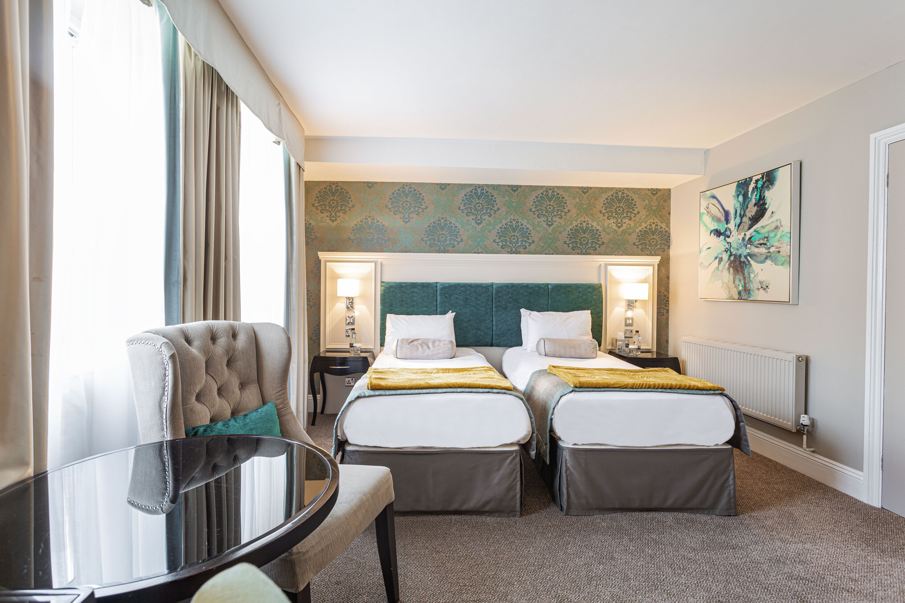Quarto de hotel com duas camas, área de estar com uma poltrona, uma mesa e uma janela grande.