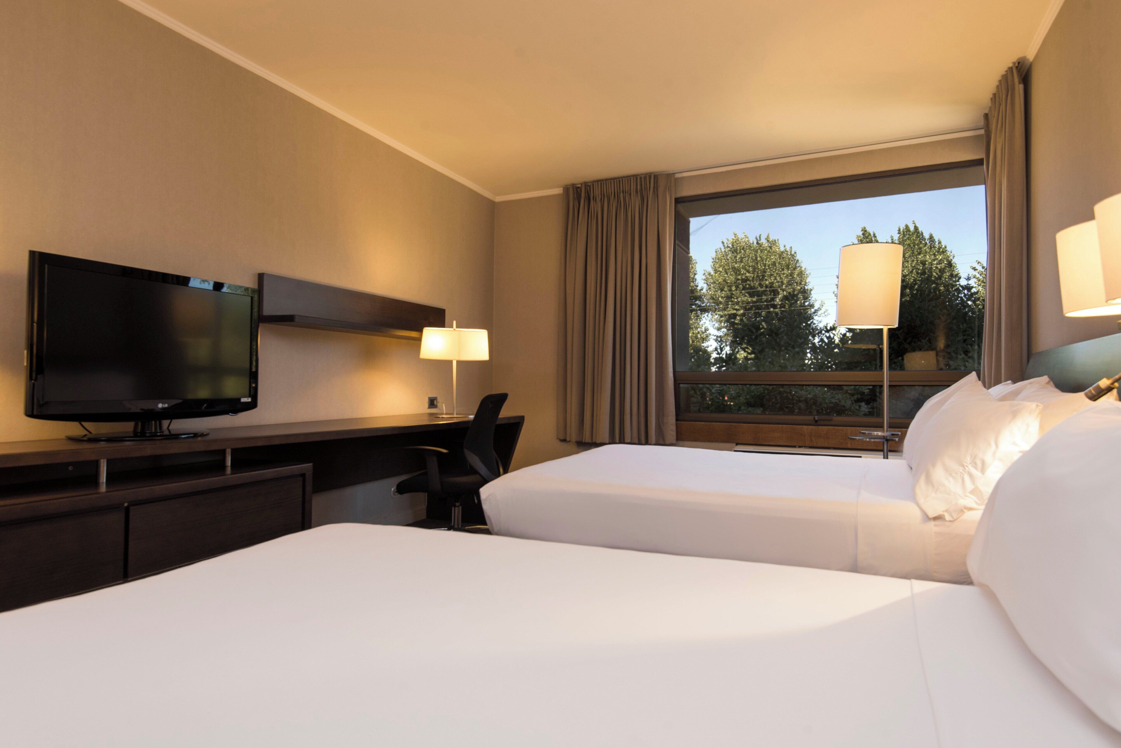 Quarto de hotel com duas camas, uma escrivaninha, televisão e janela com cortinas.