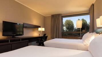 Quarto de hotel com duas camas, uma escrivaninha, televisão e janela com cortinas.