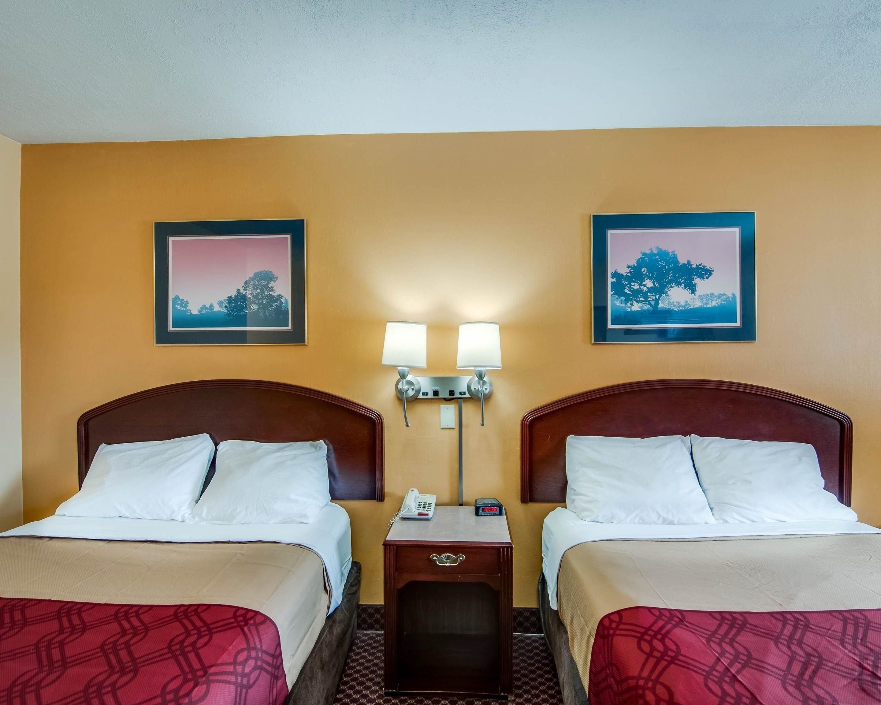 Quarto de hotel com duas camas, cada uma com roupa de cama branca e colcha estampada em vermelho. Duas luminárias fixadas na parede estão entre as camas, e duas quadros emoldurados acima delas.