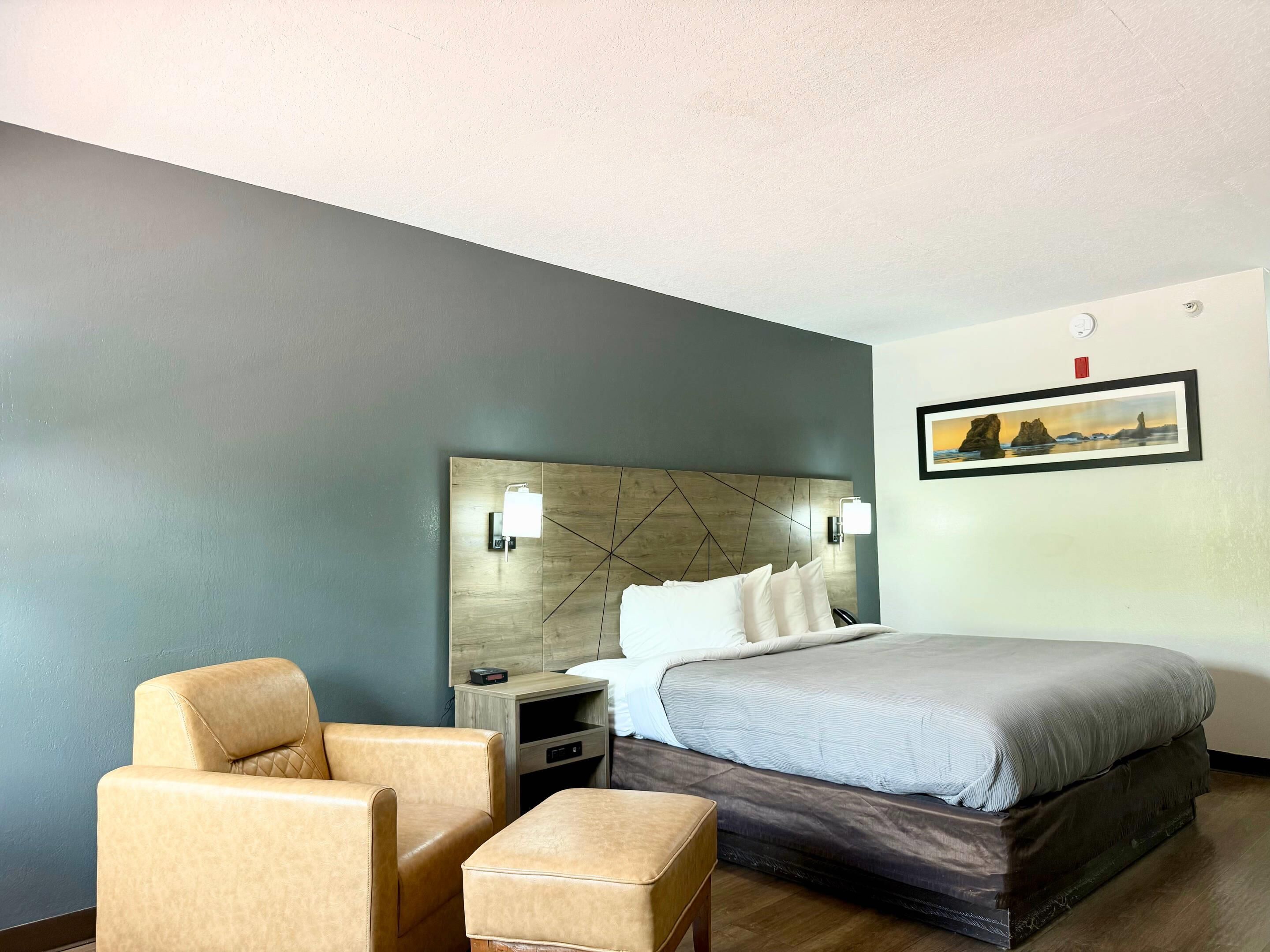 Quarto de hotel com cama, cadeira, mesa de cabeceira e um quadro na parede.