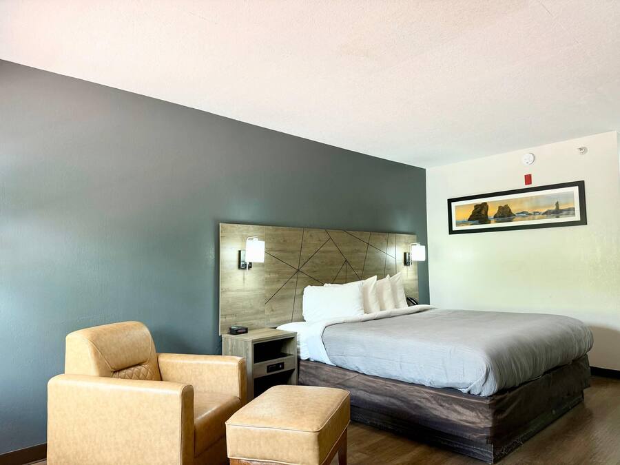 Quarto de hotel com cama, cadeira, mesa de cabeceira e um quadro na parede.