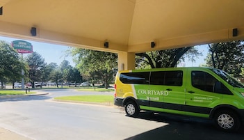 Uma van da marca Courtyard Marriott estacionada sob uma cobertura, com vista livre para árvores e uma estrada.