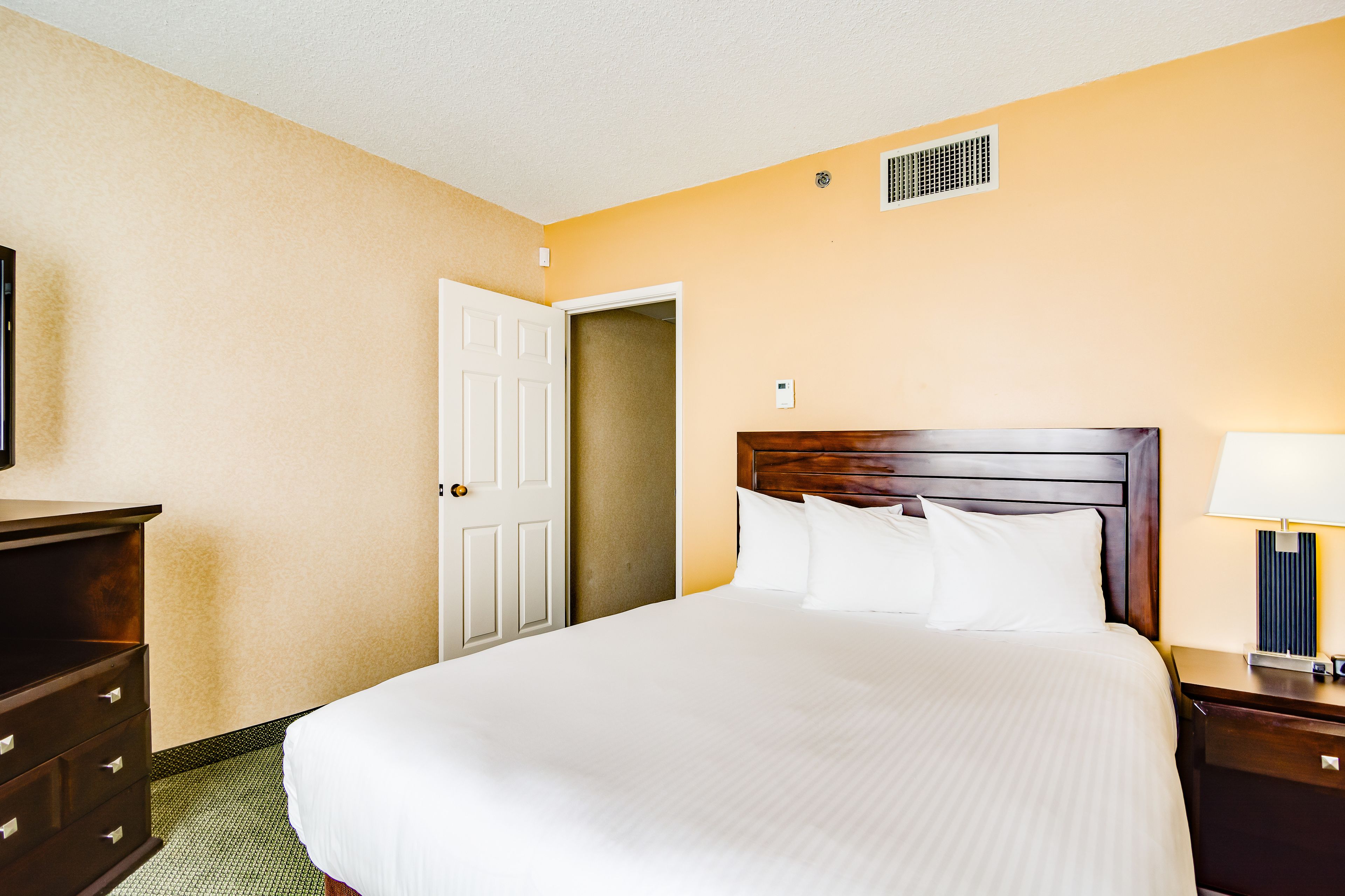 Quarto de hotel com uma cama grande, cômoda, mesa de cabeceira e luminária.
