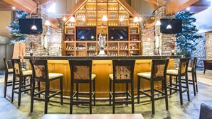 Uma área de bar com parede de pedra, prateleiras de madeira e balcão. Há seis banquetas de bar e duas televisões fixadas na parede.