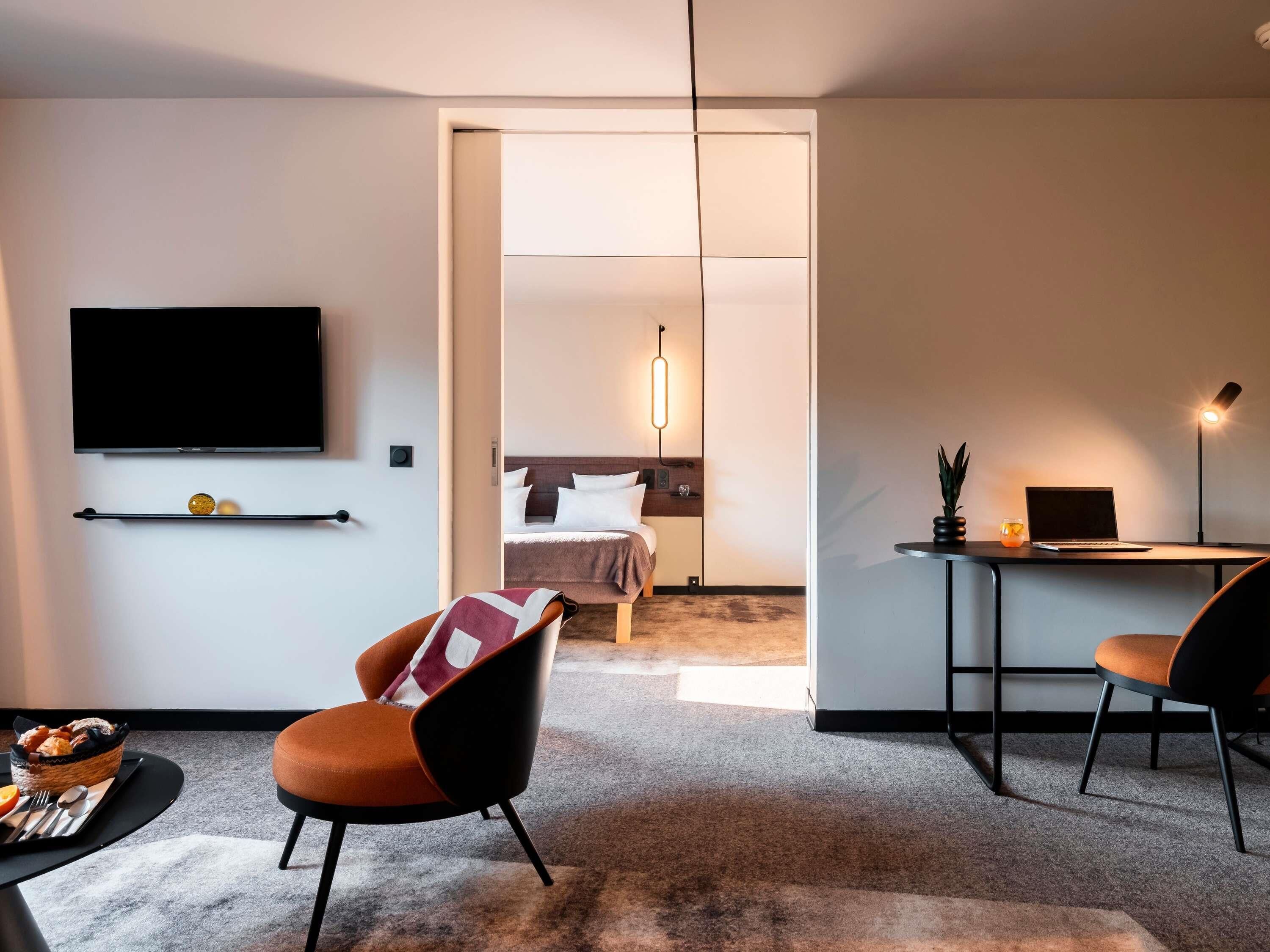 Quarto de hotel moderno com televisão de tela plana, cama com cabeceira, escrivaninha com laptop e área de refeições com cadeira e mesa.